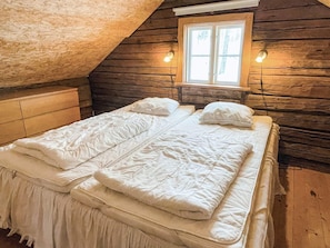 Quarto com teto inclinado de madeira, cama de casal com roupa de cama branca e duas luminárias fixadas na parede.