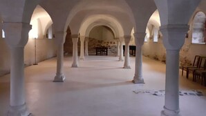 Um interior com uma série de arcos e colunas, uma mesa central e uma parede de pedra ao fundo.
