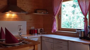 Interior de uma cabana de madeira com área de cozinha, incluindo uma mesa com uma taça de vinho, uma chaleira vermelha e uma torradeira.