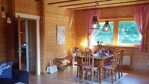 Interior acolhedor de uma cabana de madeira, com área de jantar, paredes de madeira e janela com cortinas.