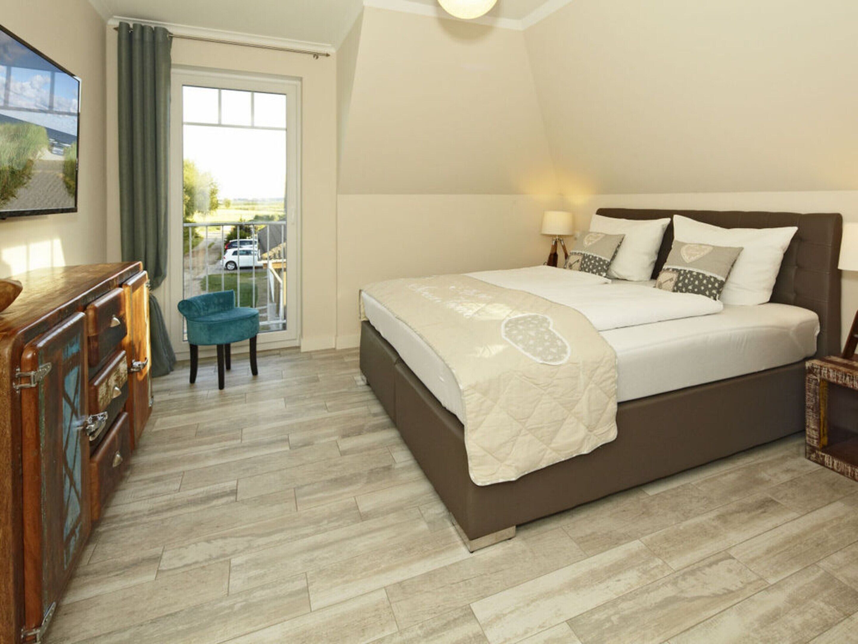 Quarto com cama grande, cômoda, cadeira e vista para o exterior pela janela.