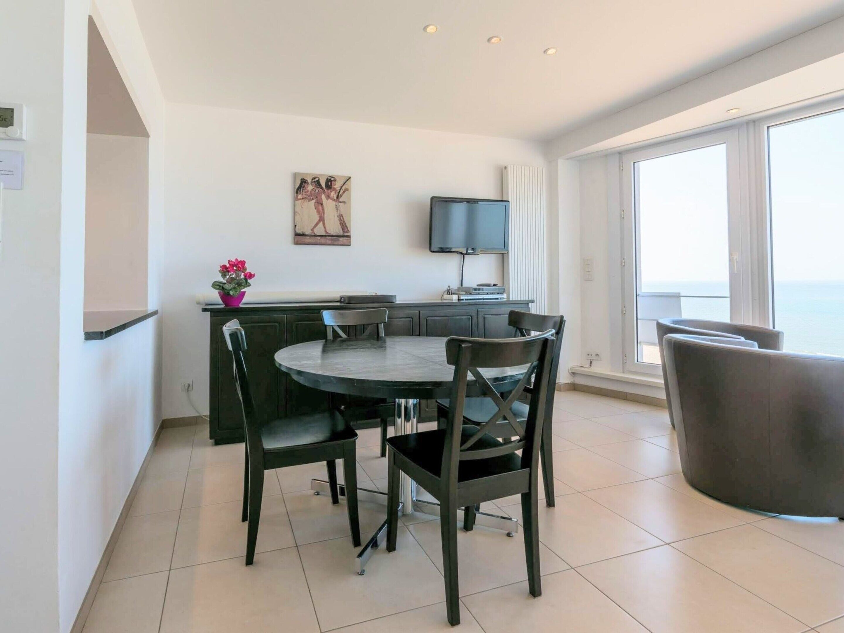 Uma sala de jantar moderna com uma mesa redonda e quatro cadeiras, uma televisão de tela plana na parede e uma grande janela com vista para o mar.