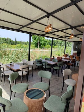Uma área de estar coberta ao ar livre, com mesas e cadeiras, uma mesa redonda com tampo de vidro e vista para um campo através de grandes janelas.