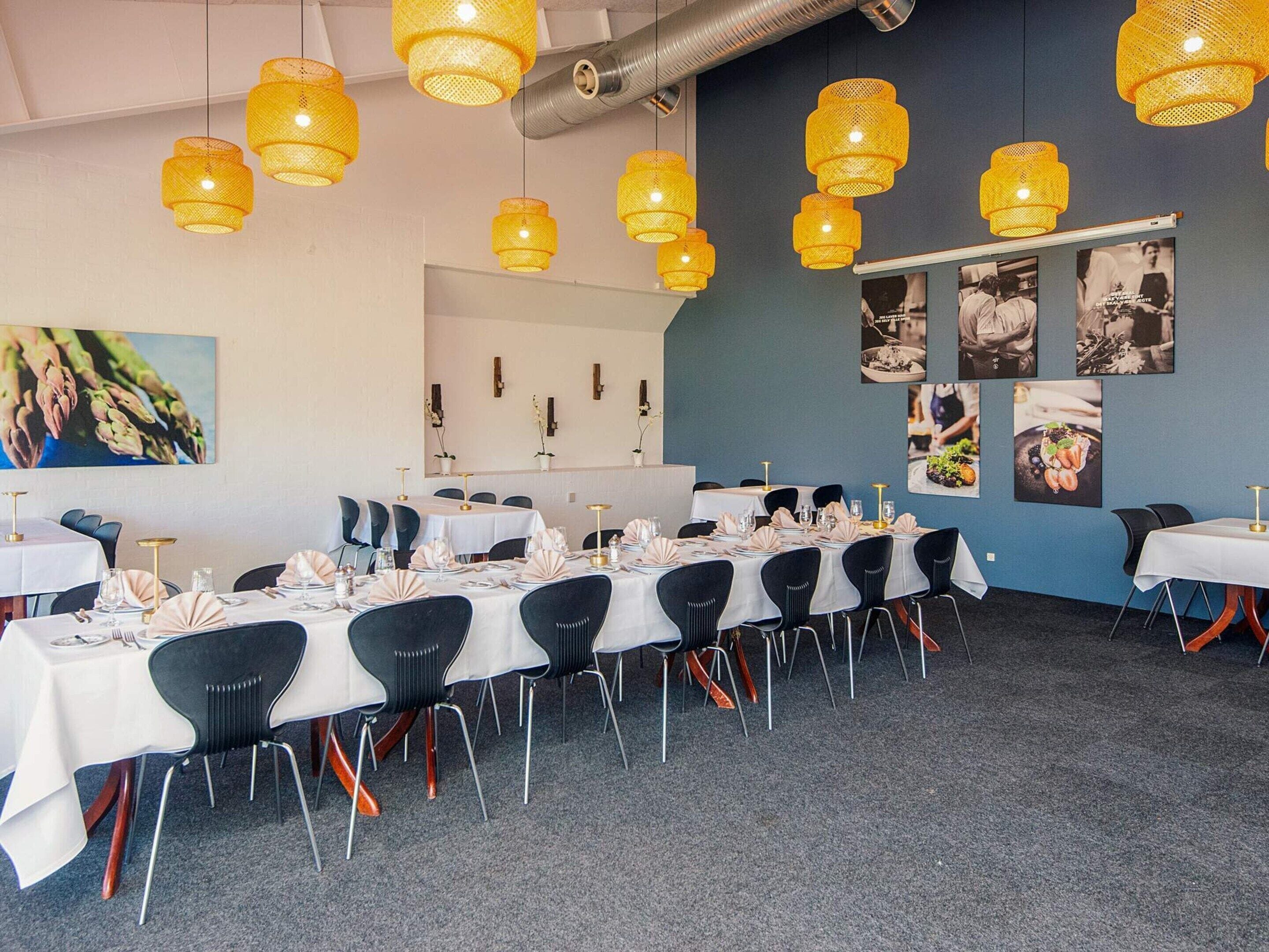 Uma sala de conferência com mesas preparadas para uma reunião, luminárias pendentes amarelas e obras de arte nas paredes.