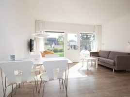 Uma sala de estar moderna com um sofá branco, uma mesa de jantar com cadeiras e uma TV fixada na parede.