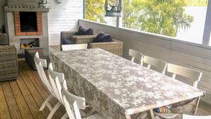 Uma área de refeições coberta ao ar livre, com uma mesa comprida, cadeiras brancas, assentos de vime e uma lareira de tijolos.