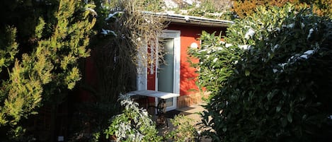 Um edifício vermelho com porta branca, rodeado por vegetação densa e plantas cobertas de neve.