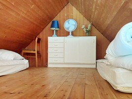 Um quarto de sótão pequeno e aconchegante, com paredes de madeira, um penteou branco com gavetas, um abajur azul, um ventilador branco e duas camas com roupa de cama branca.