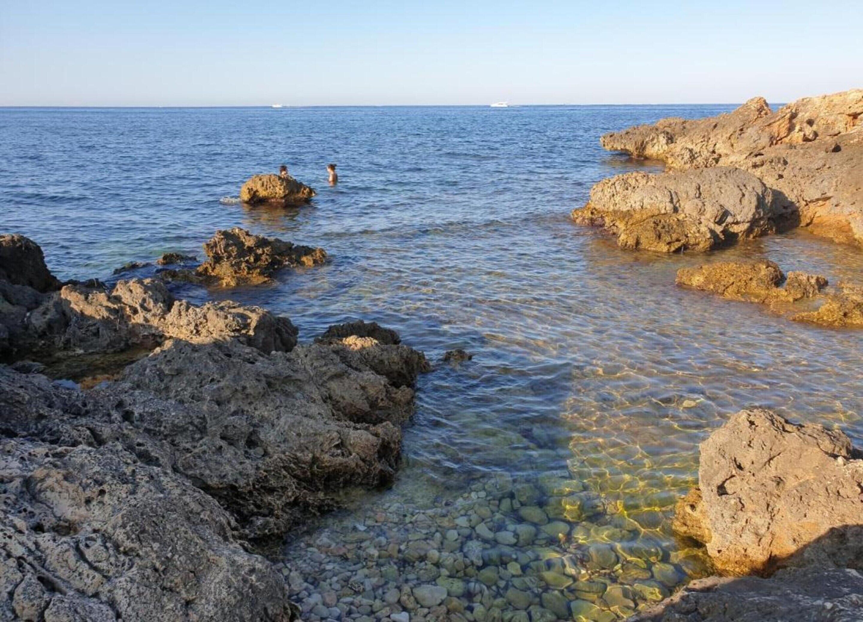 Uma costa rochosa com água límpida e barcos ao longe.