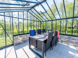 Varanda envidraçada com mesa e cadeiras, parede de tijolos e vista para o verde.