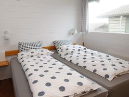 Quarto com duas camas de solteiro, cada uma com colchão branco e roupa de cama azul com bolinhas. Há um luminário fixado na parede acima das camas e uma janela com cortina à direita.