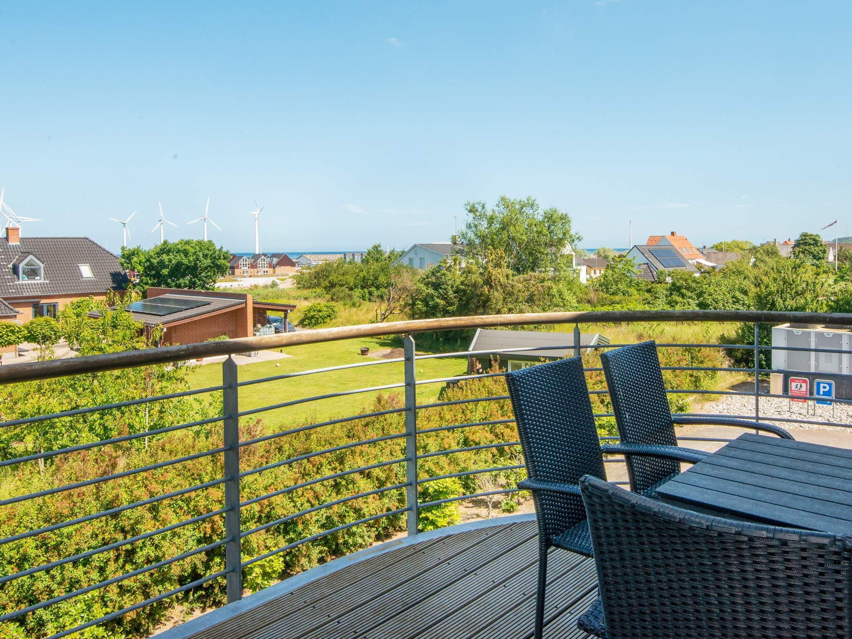 Varanda com cadeiras de vime e uma mesa, com vista para uma área residencial com casas, turbinas eólicas e um campo gramado.