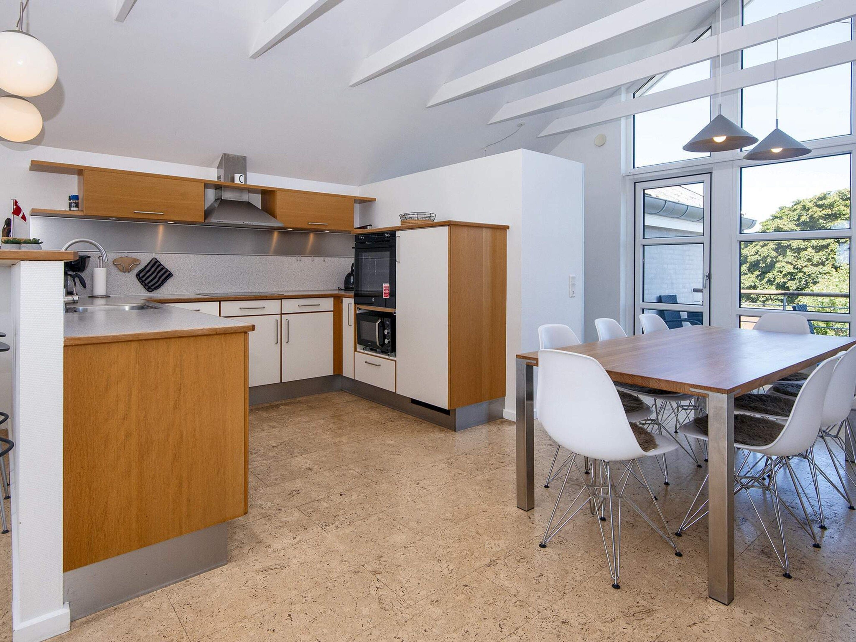 Uma cozinha moderna com ilha central, armários brancos, eletrodomésticos de aço inoxidável e uma área de refeições com cadeiras brancas.