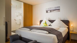 Quarto de hotel moderno com uma cama grande, um pequeno espaço de estar, banheiro com porta de vidro e um quadro na parede.