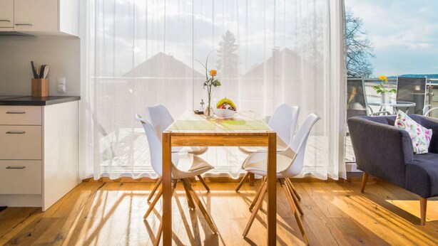 Uma sala de jantar com mesa posta para dois, um sofá e vista para o exterior por uma grande janela.