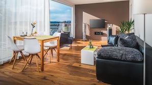 Uma sala de estar moderna com piso de madeira, um sofá preto, uma mesa de jantar com cadeiras e uma grande janela com vista para o céu e prédios.