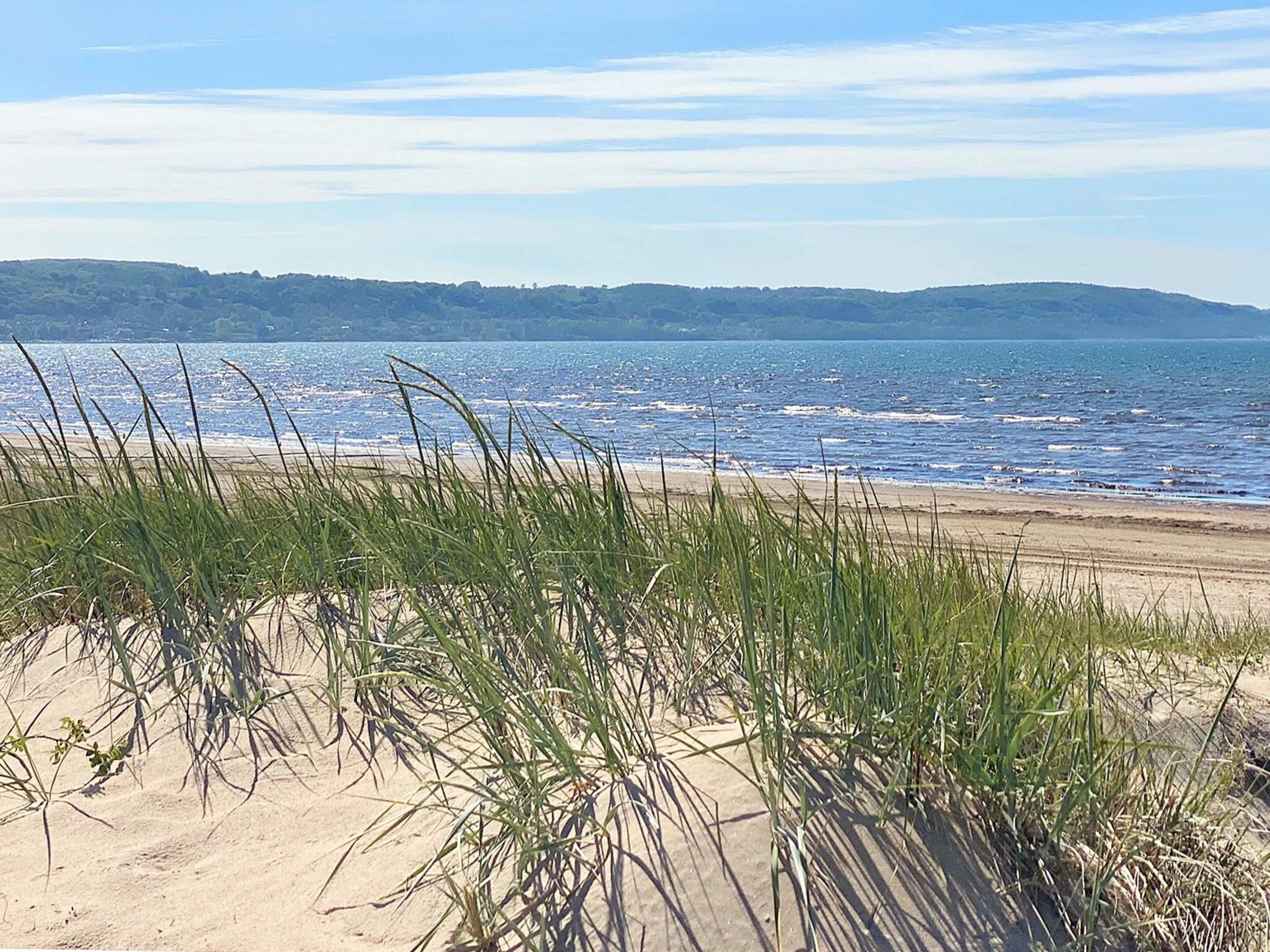 Uma praia com dunas de areia e grama ao primeiro plano, que se estende até um mar calmo e um litoral distante.