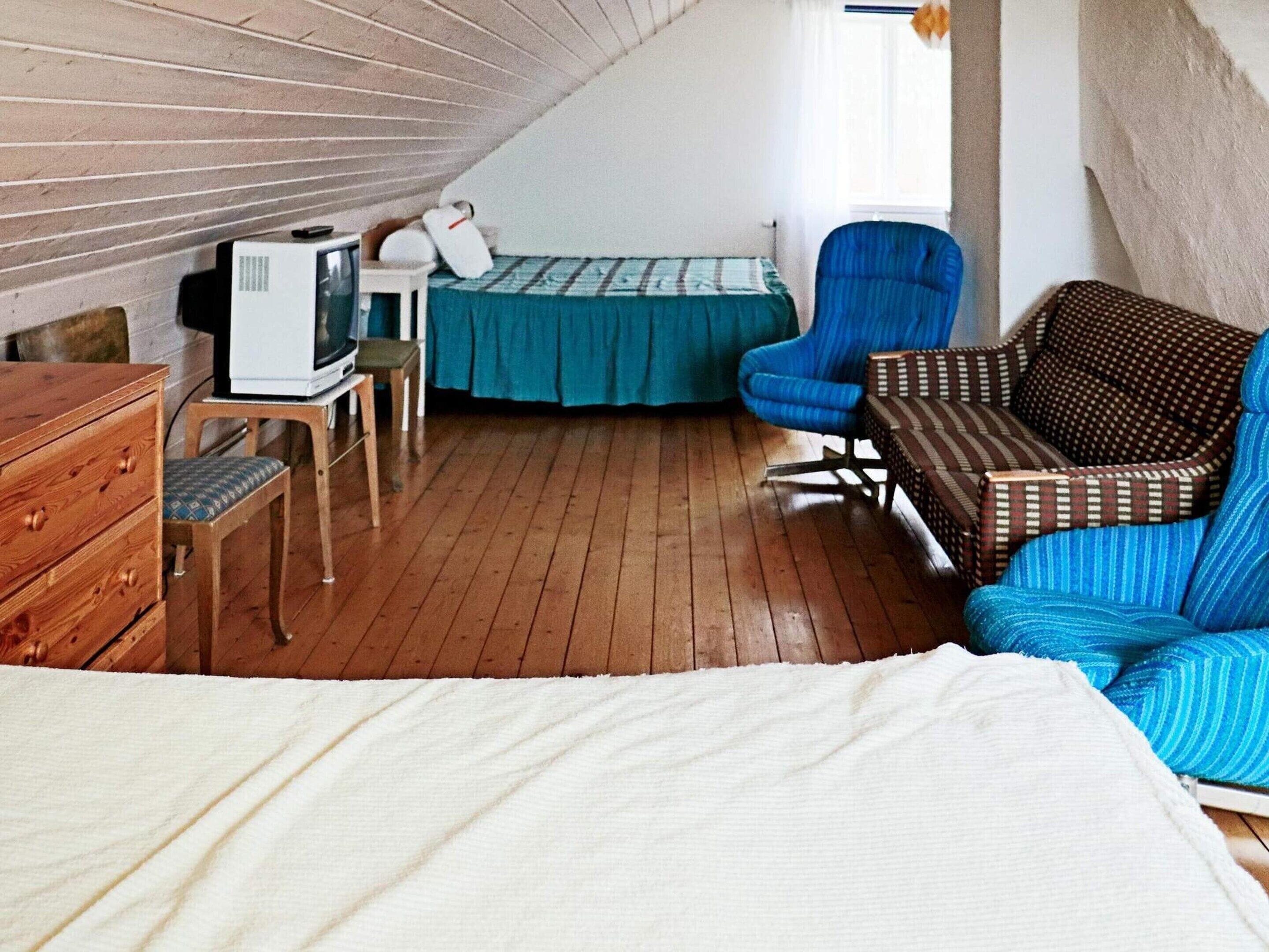Quarto compacto com piso de madeira, cama com cobertura branca, poltrona azul, sofá de vime, mesinha com televisão e cômoda de madeira.