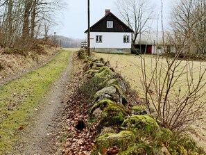 Uma estrada rural que leva a uma casa branca com telhado escuro, cercada por um muro de pedra e árvores.
