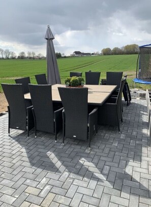 Um pátio externo com mesa e cadeiras, uma planta em vaso e vista para um campo verde.