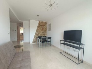 Uma sala de estar com sofá, televisão em um suporte, área de jantar com cadeiras, escada e um lustre.