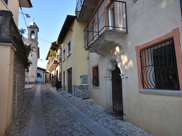 Uma rua de paralelepípedos estreita, ladeada por edifícios com varandas e uma torre de sino ao fundo.