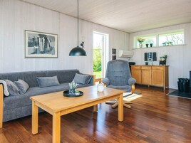 Uma sala de estar moderna com um sofá cinzento, uma mesa de centro de madeira e uma poltrona cinza. Há um armário de madeira com uma TV e uma janela com plantas.