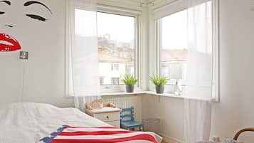 Quarto com cama, cadeira, mesa pequena, duas janelas com cortinas e vista para prédios.