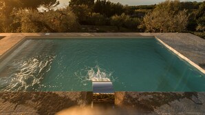 Piscina retangular com um elemento aquático no centro, rodeada por degraus de pedra e vista para um lago e colinas.