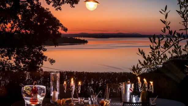 Mesa posta para refeição, com velas e garrafa, vista para um lago calmo ao pôr do sol.