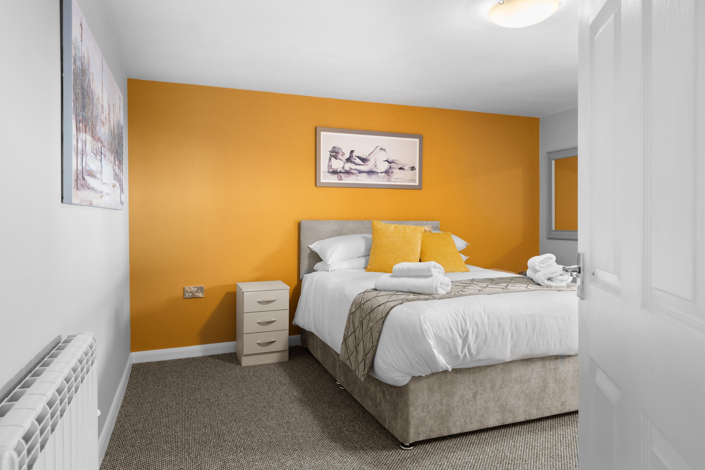 Quarto com cama, mesa de cabeceira, radiador e quadro na parede.