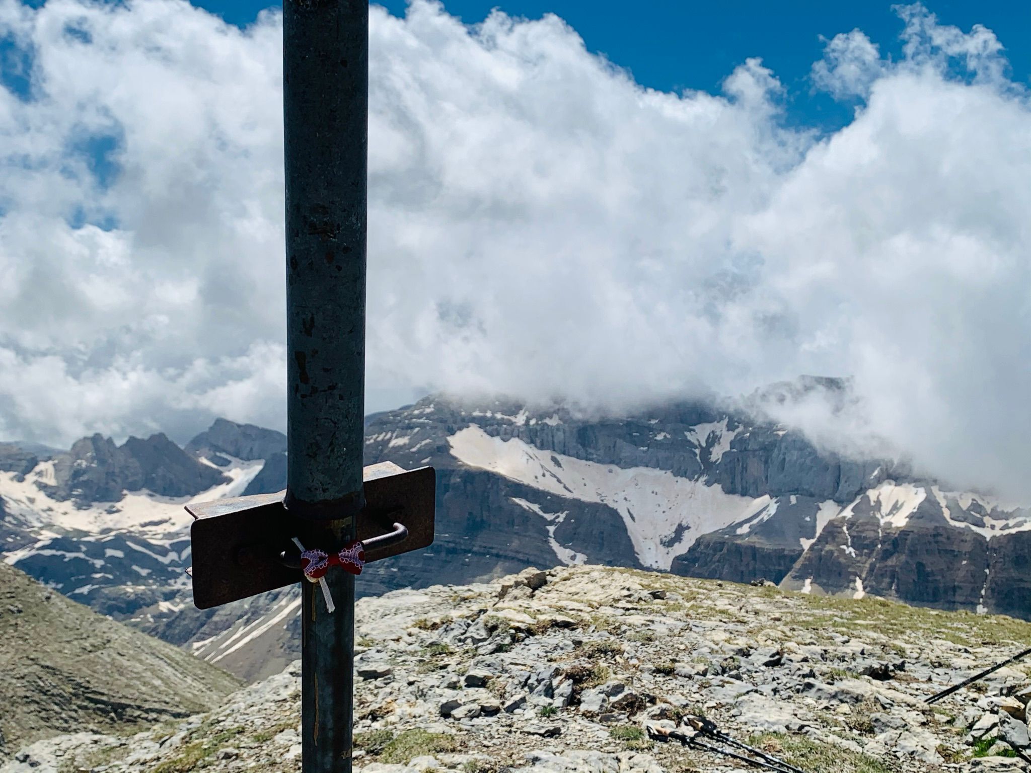 Um pico de montanha com um poste de metal e uma placa, rodeado por montanhas nevadas e nuvens.