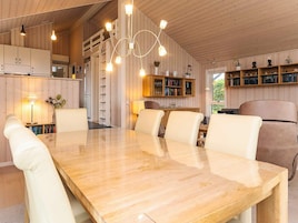 Uma sala de jantar ampla com uma mesa e cadeiras de madeira, uma cozinha ao fundo e uma estante repleta de livros.
