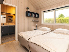 Quarto com duas camas, teto de madeira, janela com vista para o verde, uma estante e uma área de cozinha visível por uma porta aberta.