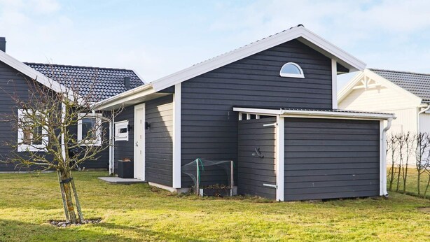 Uma casa moderna térrea, com fachada escura, porta de garagem branca e um pequeno jardim frontal com uma árvore.