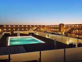 Uma área de piscina no terraço, com uma piscina retangular, rodeada por um edifício com várias andares, varandas e uma galeria coberta.