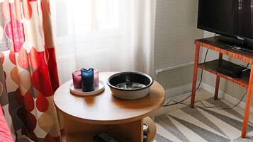 Quarto com televisão sobre suporte de madeira, mesa redonda com tigela e velas, controle remoto e tapete estampado.