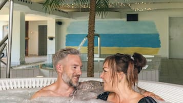 Um casal em uma banheira de hidromassagem, com uma palmeira e um muro azul e amarelo ao fundo.