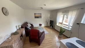 Sala de estar com sofá, mesa de centro e televisão. Há uma janela com cortinas e uma planta no chão.