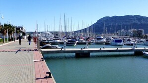 Uma marina com barcos atracados, um calçadão pavimentado e uma montanha ao fundo.