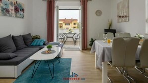 Uma sala de estar moderna com sofá, mesa de centro e área de jantar. Há uma varanda com vista para edifícios.