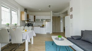Uma cozinha moderna com armários brancos, uma sala de jantar com mesa posta para dois e uma sala de estar com sofá e mesa de centro.