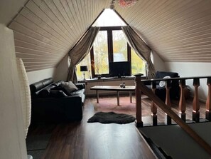 Um quarto de sótão aconchegante com piso de madeira, um sofá de couro preto, uma mesinha e uma televisão.