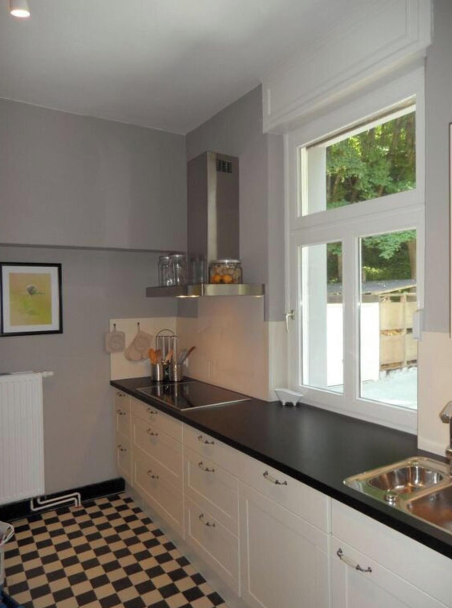 Uma cozinha com piso xadrez preto e branco, armários brancos, bancada preta e uma janela com vista para o verde lá fora.