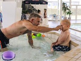 Um homem e uma criança numa área de piscina, com um muro de pedra e um guarda-sol com a marca Pepsi.