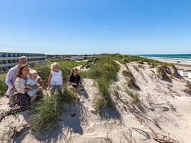 Uma família em uma praia arenosa, com dunas e um edifício ao fundo.