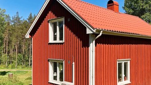 Uma casa de madeira vermelha com telhado inclinado, duas janelas e um aparelho de ar condicionado na lateral.