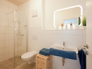 Um banheiro moderno com chuveiro de vidro, azulejos brancos, vaso sanitário, pia com toalha azul e espelho com moldura iluminada.