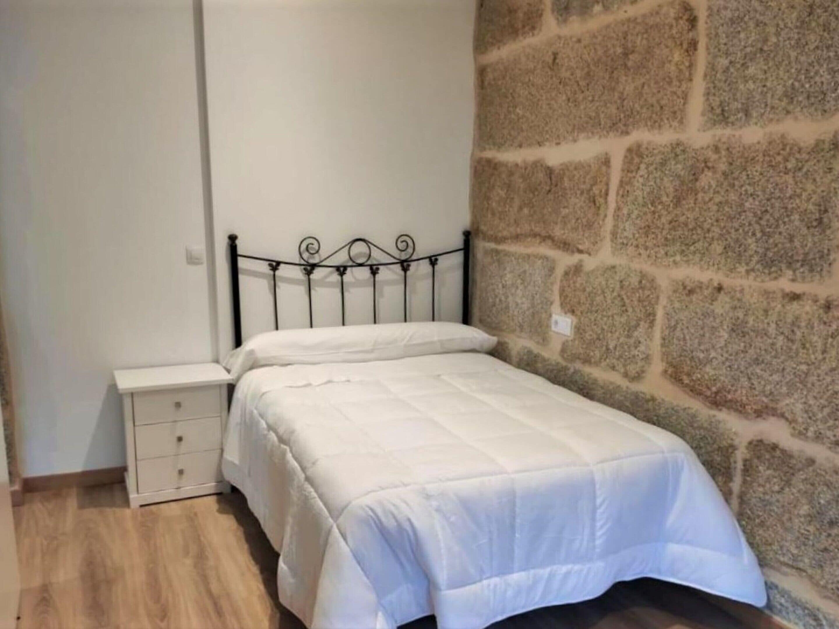 Uma cama de solteiro com cabeceira de metal em quarto com parede de pedra e chão de madeira.