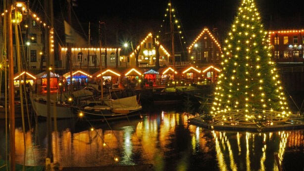 Uma área à beira-mar com edifícios decorados com luzes de corda, uma grande árvore de Natal iluminada e barcos atracados na água.